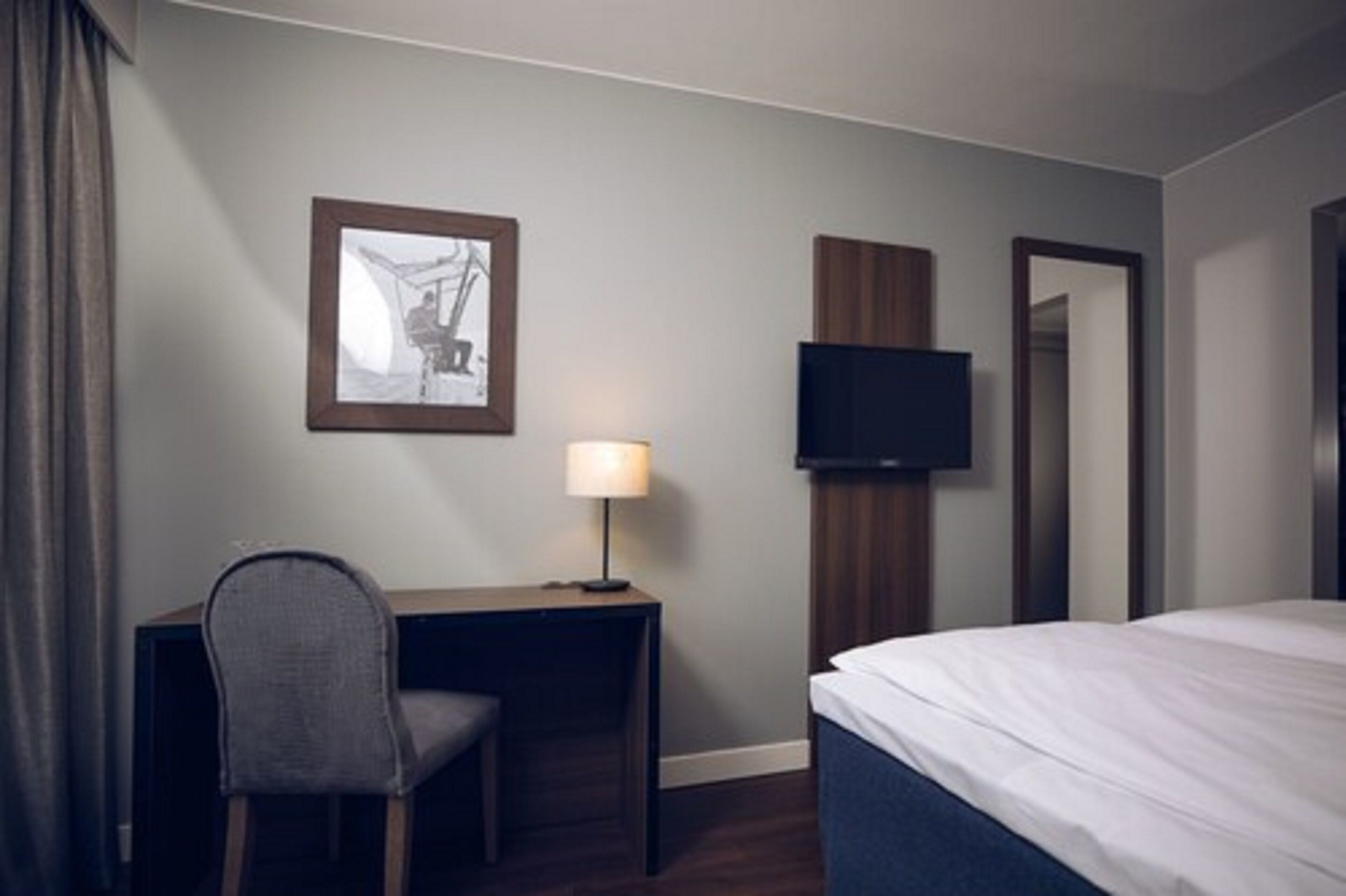 Quarto de hotel com cama, escrivaninha com cadeira, luminária, televisão e um quadro na parede.