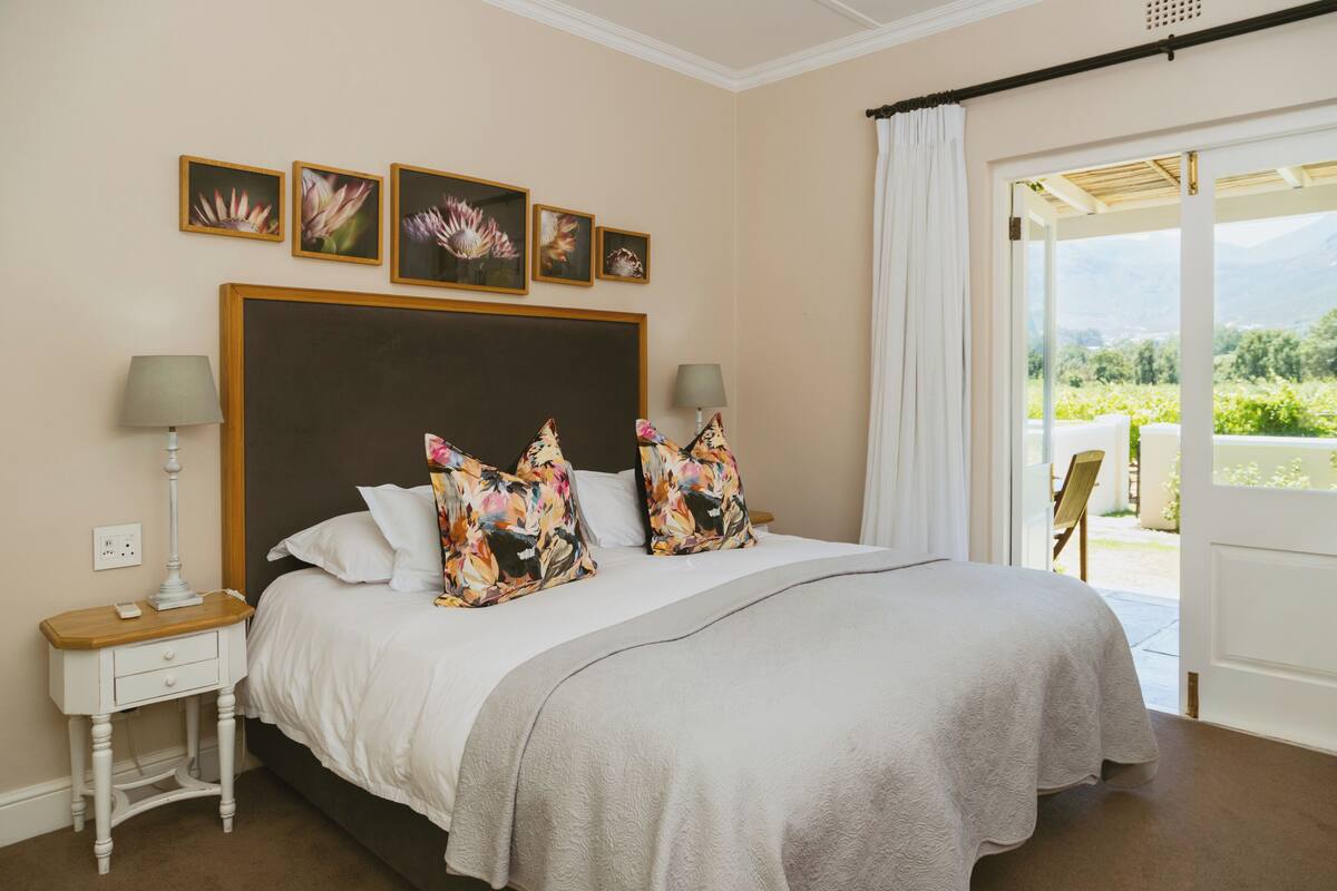 Quarto com uma cama grande, almofadas florais, uma mesa de cabeceira e vista para o exterior.