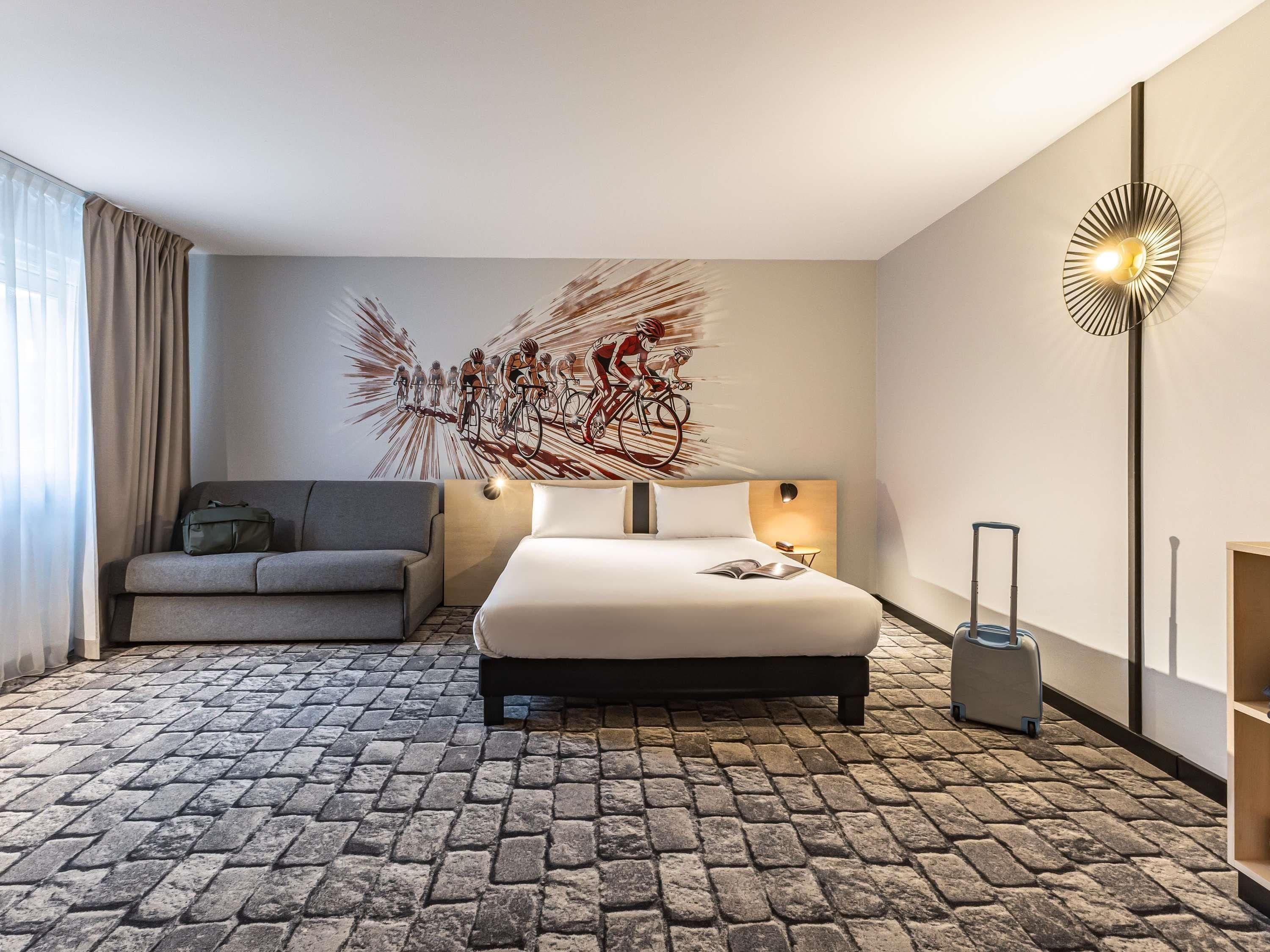 Quarto de hotel com cama, sofá, painel na parede, abajur e mala.