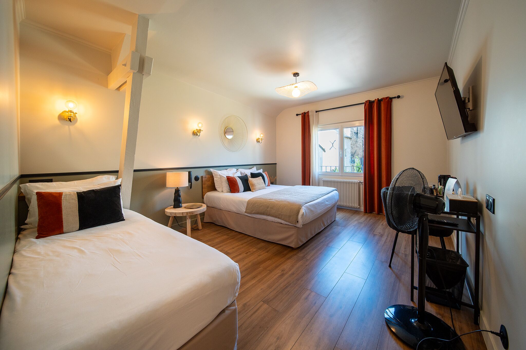 Quarto de hotel com duas camas, uma mesa pequena, uma cadeira, um ventilador e uma TV fixada na parede.