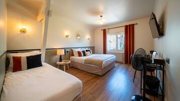 Quarto de hotel com duas camas, uma mesa pequena, uma cadeira, um ventilador e uma TV fixada na parede.