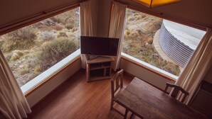 Quarto com televisão em suporte de madeira, mesa de jantar e duas cadeiras. Há janelas amplas com cortinas, e um abajur amarelo pendurado no teto.