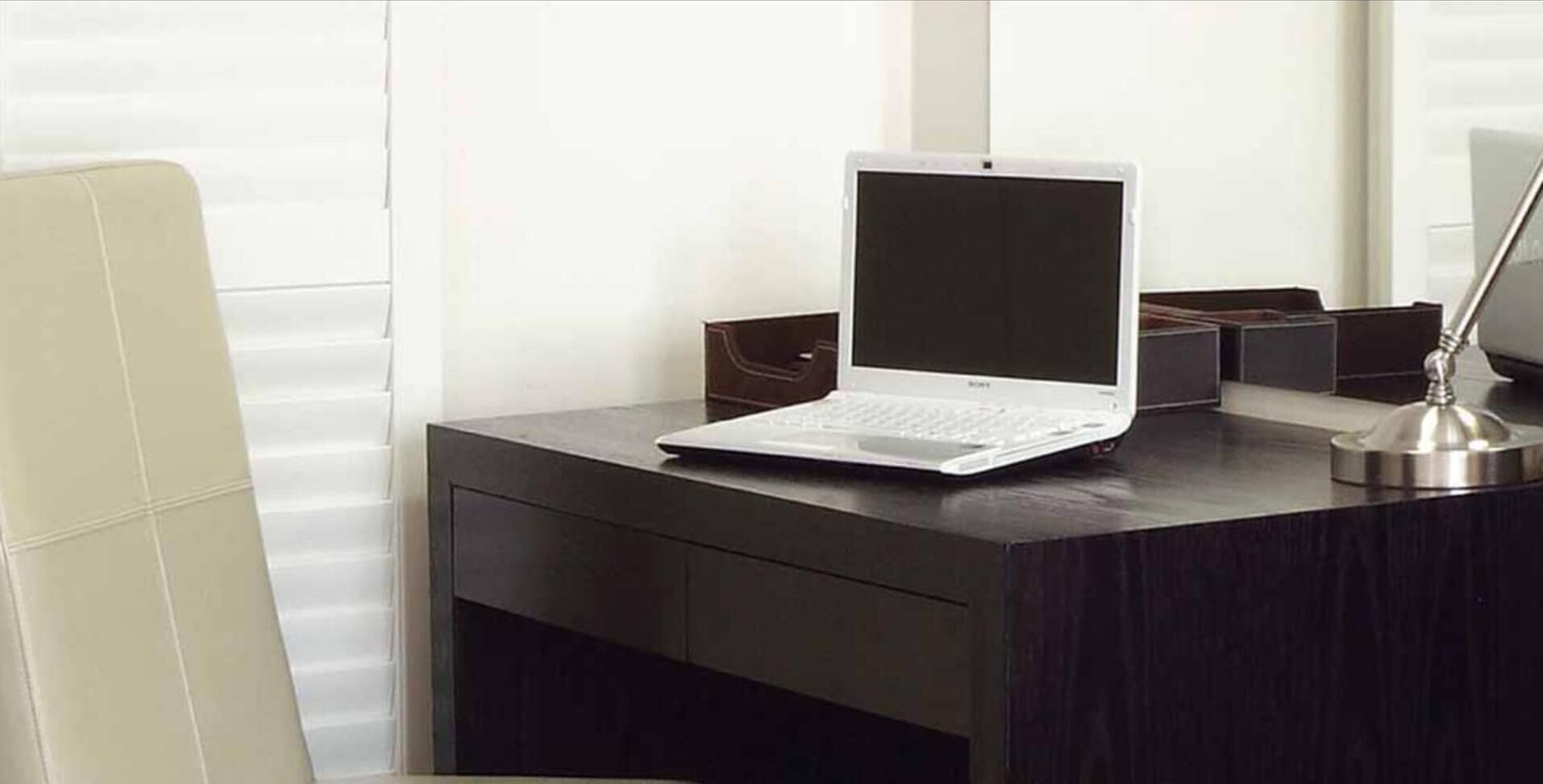 Um laptop sobre uma escrivaninha de madeira escura com uma gaveta, um objeto marrom pequeno e uma luminária de mesa.