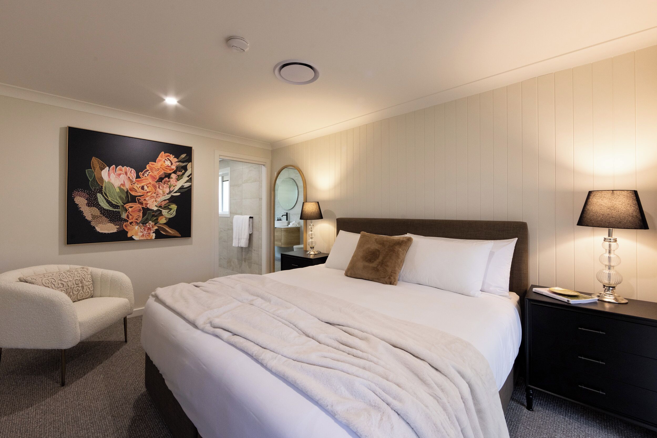 Quarto de hotel com uma cama grande, uma cadeira, mesa de cabeceira com abajur, espelho e um quadro na parede.