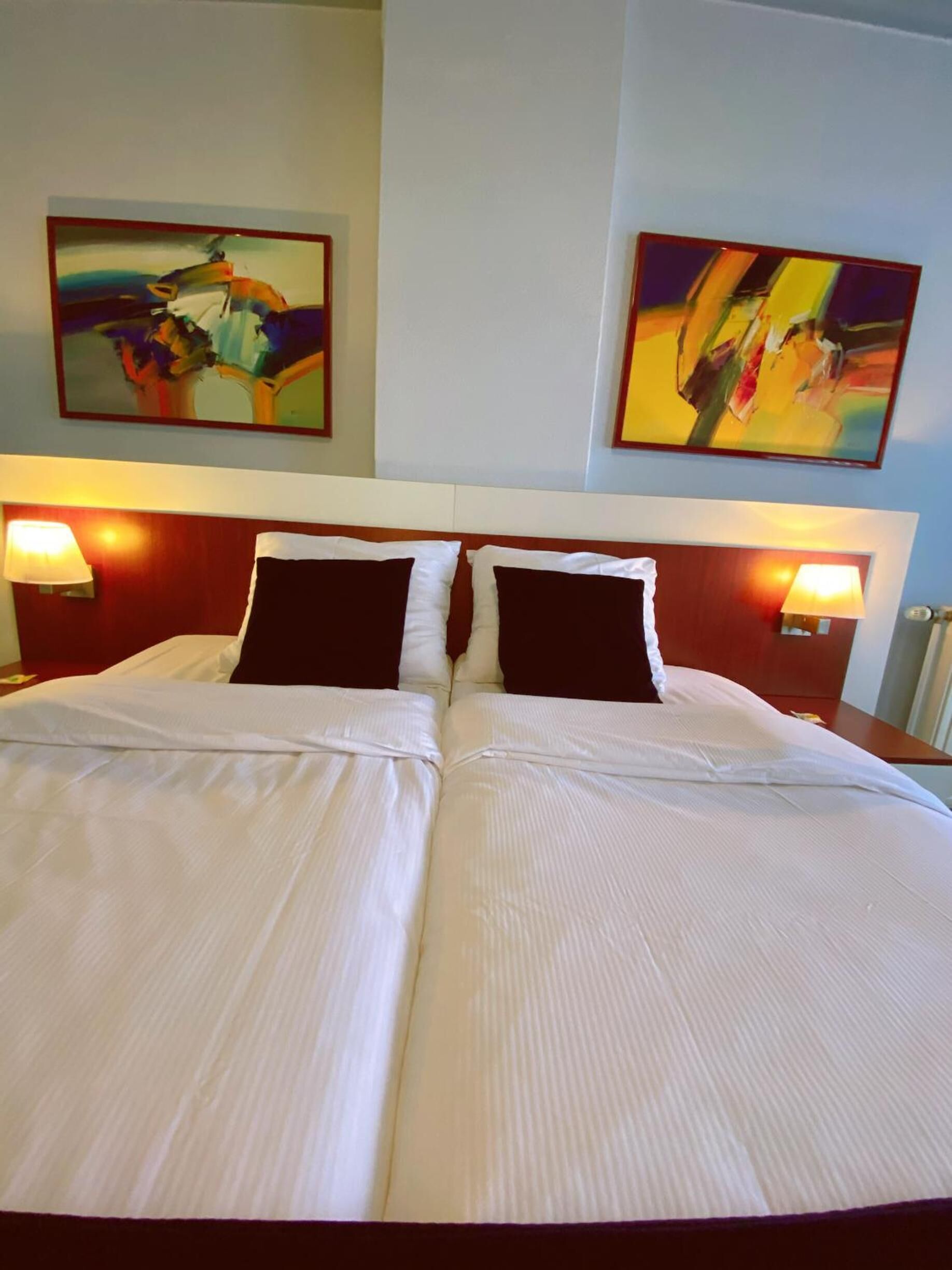 Uma cama de casal com roupa de cama branca, duas almofadas escuras e duas luminárias fixadas na parede.