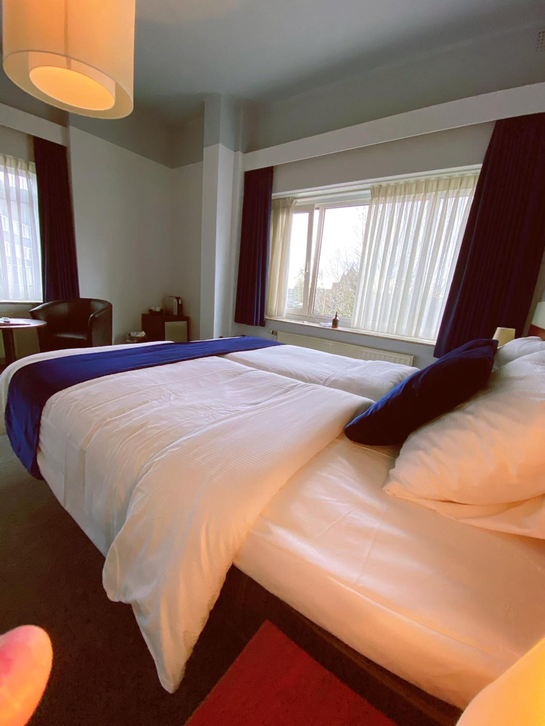 Uma cama de casal com roupa de cama branca e azul, uma escrivaninha com cadeira, uma janela com cortinas e um tapete vermelho.