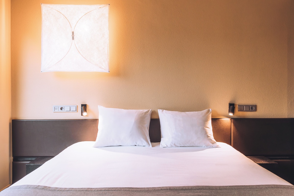 Quarto de hotel com uma cama grande, duas mesas de cabeceira, uma luminária fixada na parede e uma tomada.
