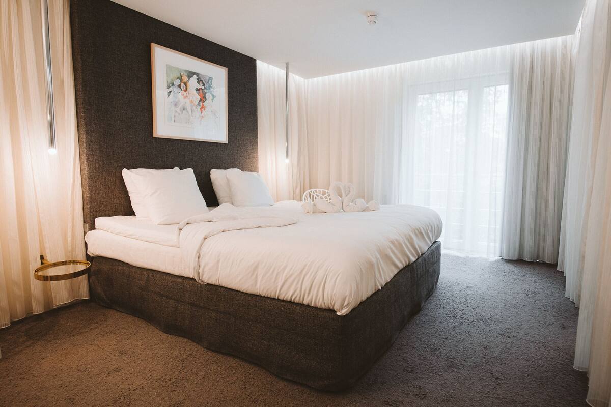 Quarto moderno com cama grande, roupa de cama branca, cabeceira escura, uma obra de arte emoldurada na parede e cortinas leves.