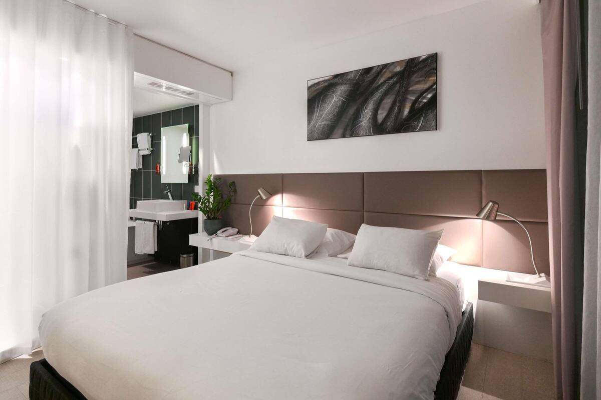 Quarto de hotel moderno com uma cama grande, mesas de cabeceira, um espelho e uma obra de arte na parede.