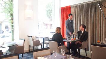 Uma sala de jantar de hotel com mesas preparadas para quatro pessoas, um garçom servindo comida e vista para a rua através de grandes janelas.