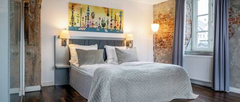 Quarto com cama, cabeceira, quadro, janela com cortinas e parede de tijolos.