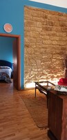 Quarto com parede de pedra, piso de madeira, cama com roupa de cama estampada, mesa com tampo de vidro e almofada vermelha.