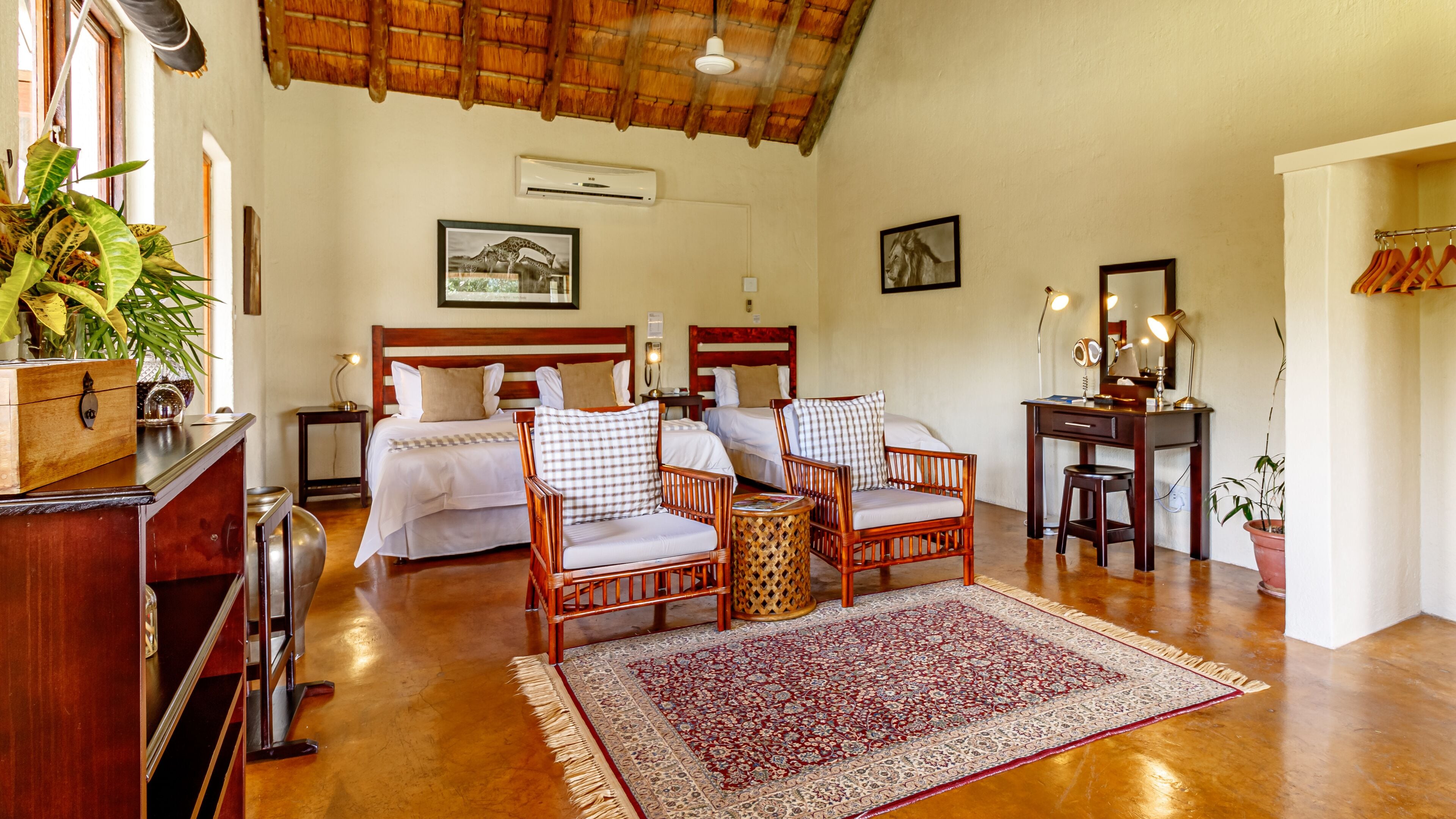 Quarto de hotel com duas camas, uma escrivaninha, uma cadeira, uma mesinha, um espelho e uma janela grande.