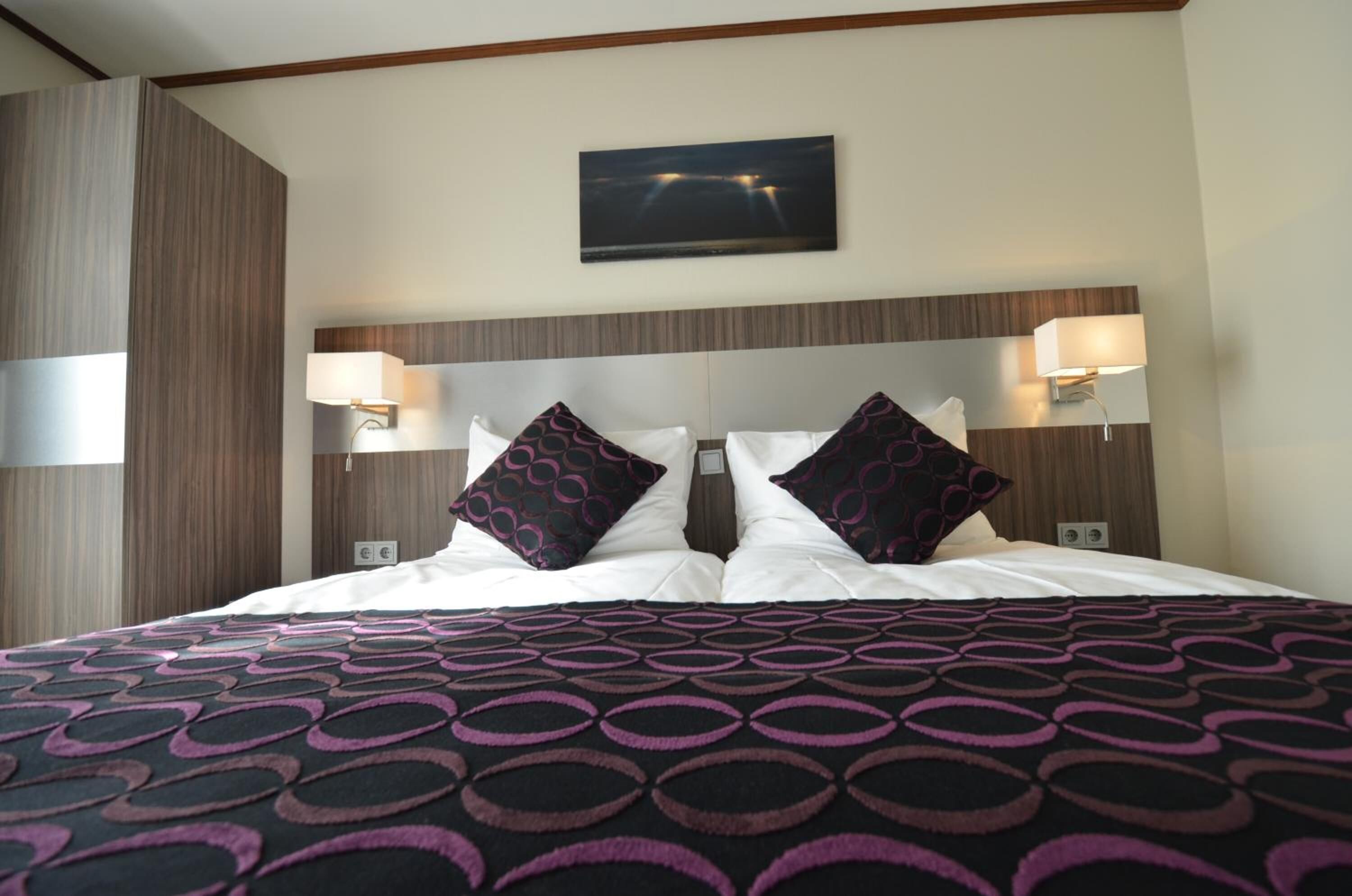 Quarto de hotel com uma cama grande, duas luminárias fixadas na parede e um quadro emoldurado sobre a cama.