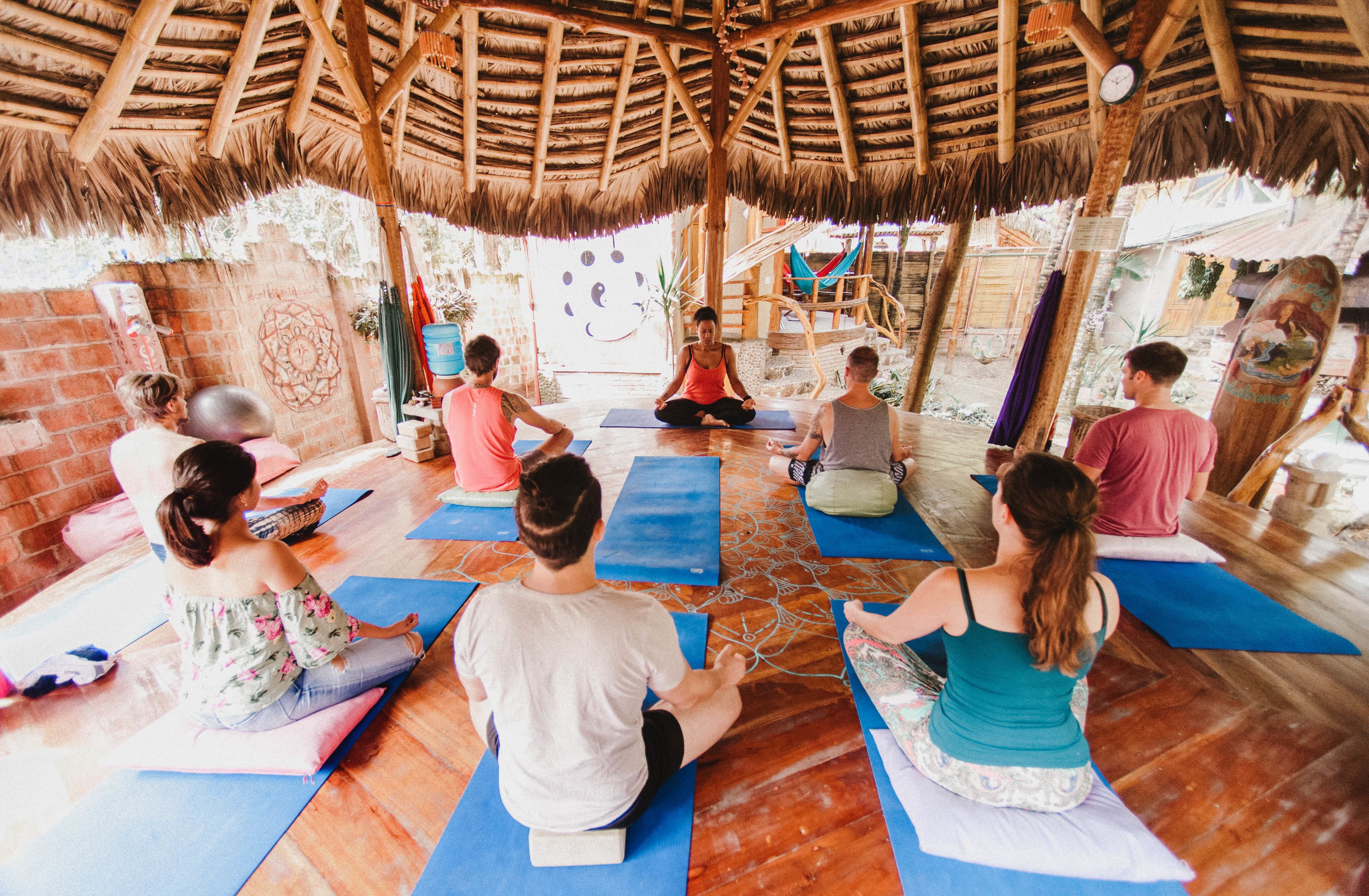 Um grupo de pessoas sentadas em tapetes de yoga num espaço com telhado de palha e paredes de tijolo.