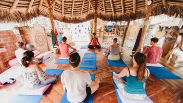 Um grupo de pessoas sentadas em tapetes de yoga num espaço com telhado de palha e paredes de tijolo.