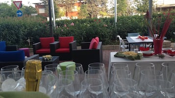 Uma área de refeições ao ar livre com móveis de vime, uma mesa posta com taças de vinho e vista para um jardim com um arbusto de flores vermelhas.