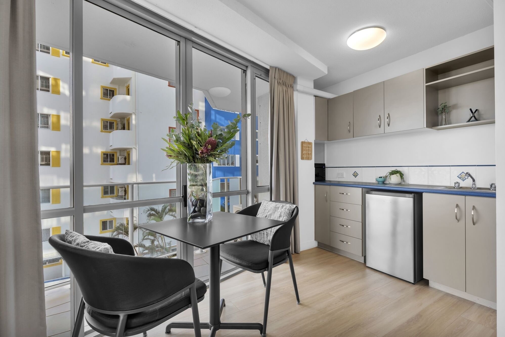 Cozinha moderna com área de refeições, janelas amplas e vista para um edifício com persianas amarelas.