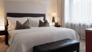 Quarto de hotel com uma cama grande, mesa de cabeceira com abajur e janela com cortinas leves.
