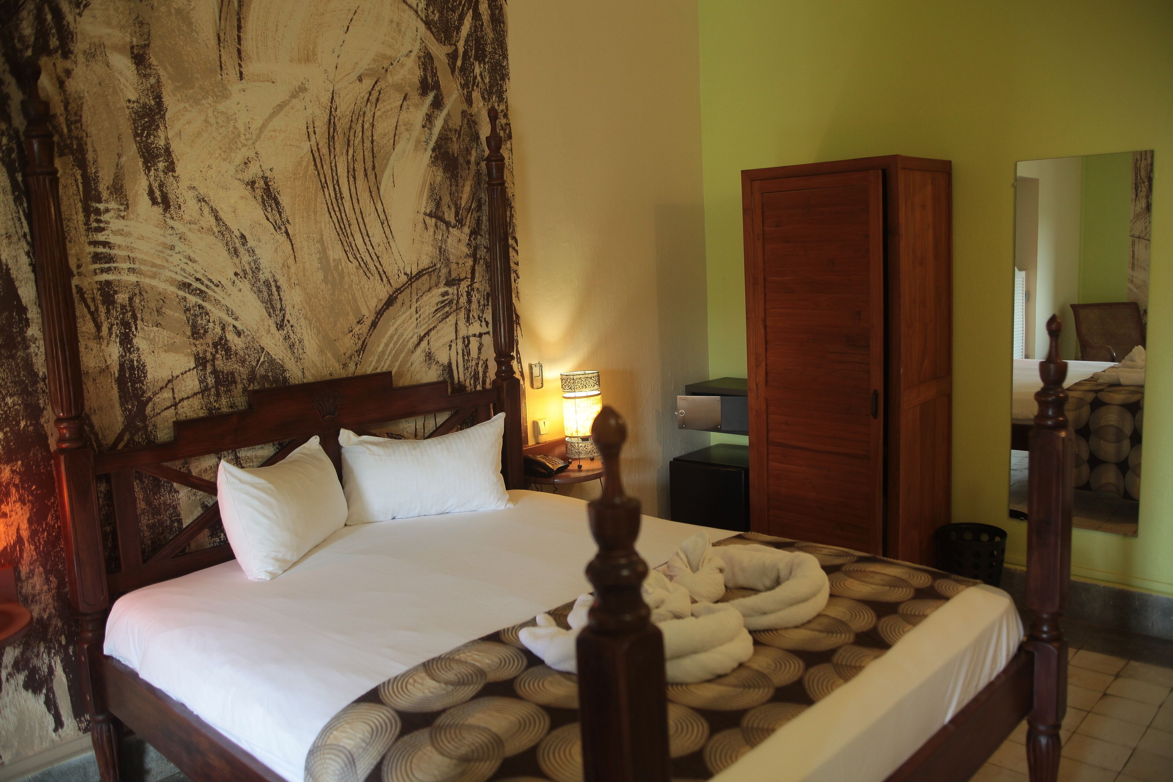 Cama bem arrumada com lençóis brancos e colcha estampada, mesa de cabeceira de madeira com luminária e guarda-roupa de madeira grande.