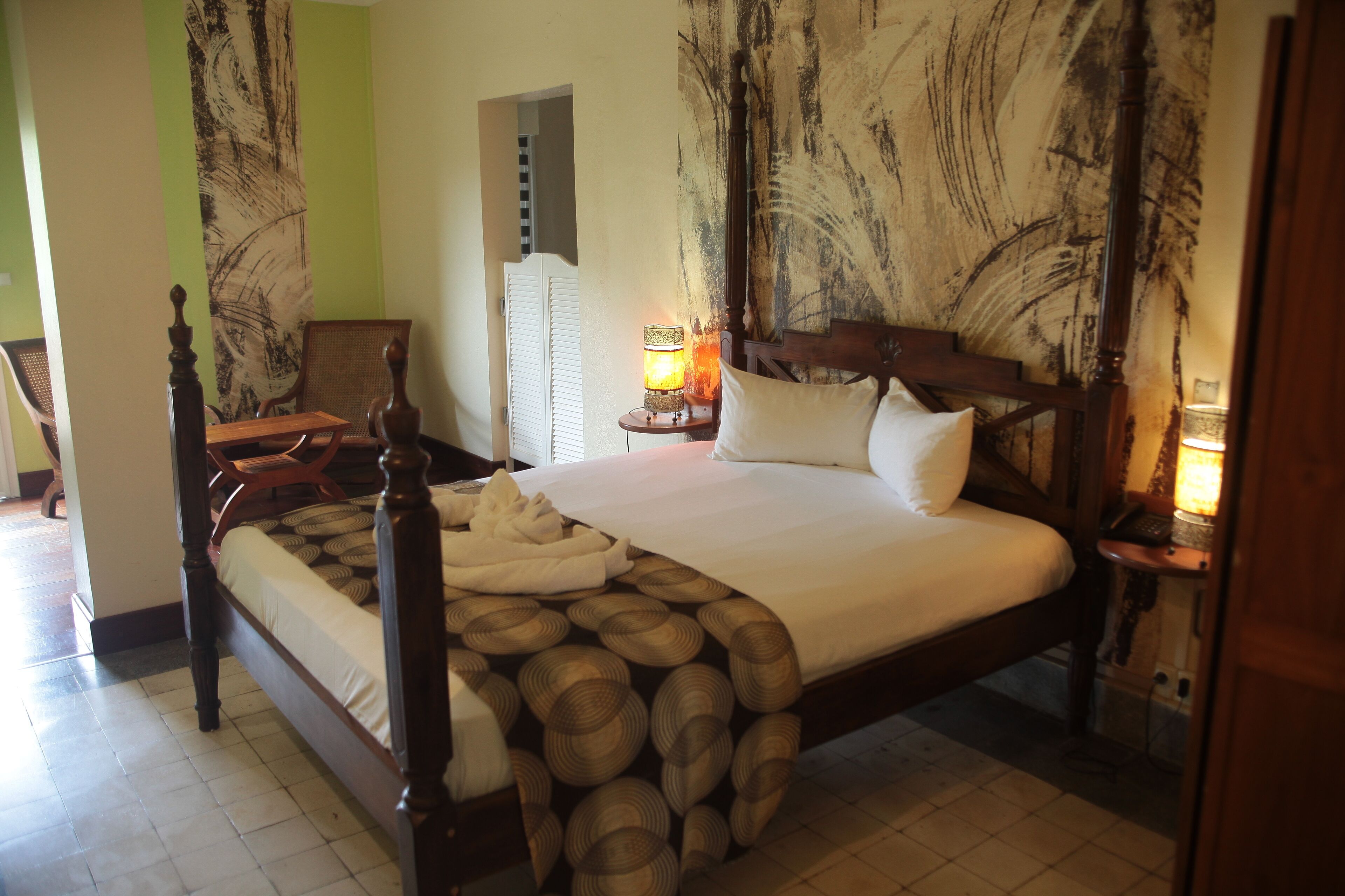 Quarto com cama de madeira, uma cadeira, mesa de cabeceira com abajur e parede com arte abstrata.