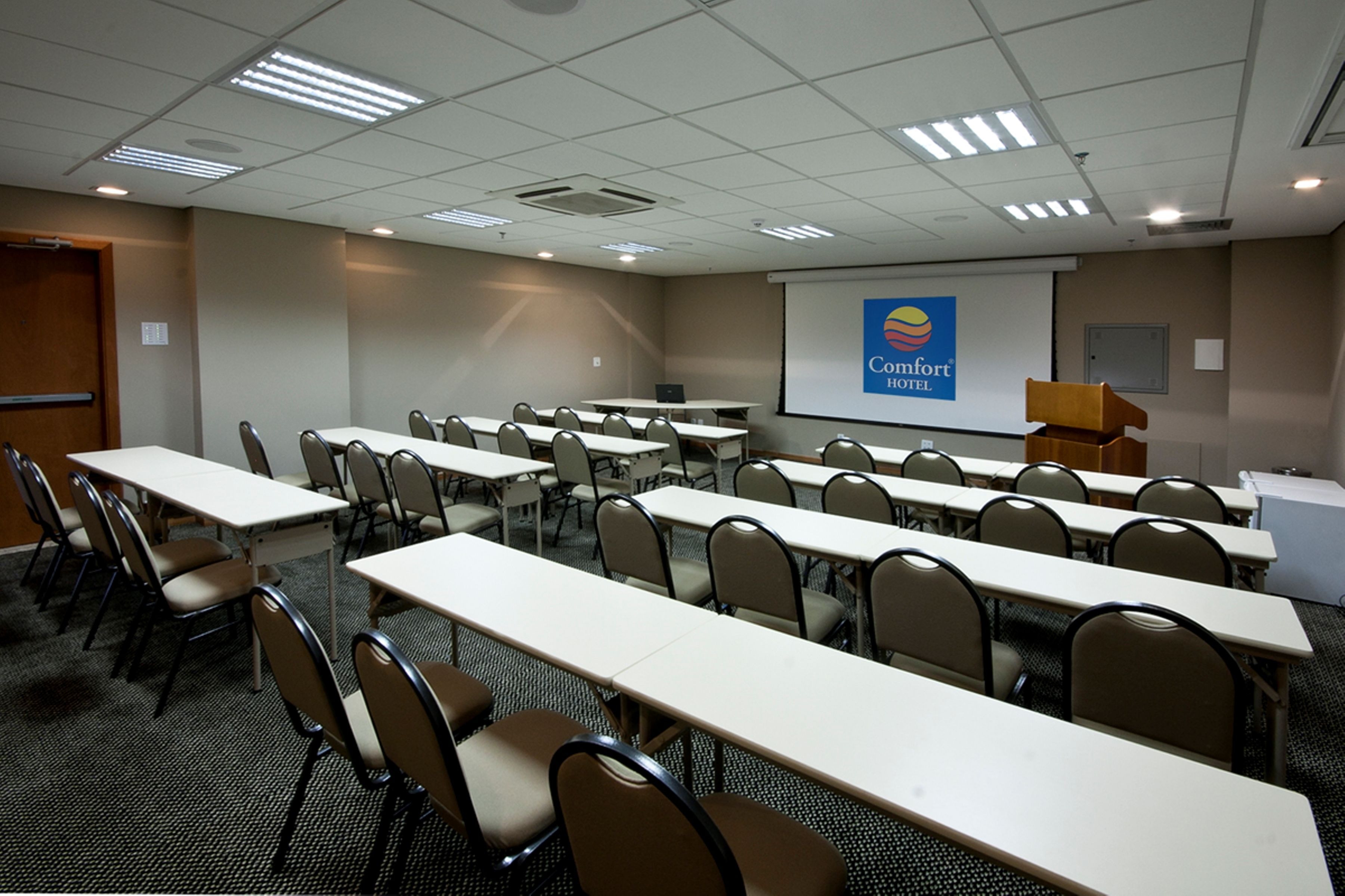 Uma sala de conferências com várias mesas e cadeiras, uma tela de projeção exibindo o logotipo do Comfort Hotel e um pódio.
