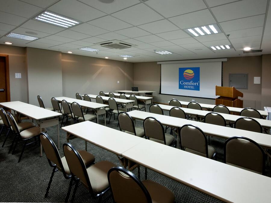 Uma sala de conferências com várias mesas e cadeiras, uma tela de projeção exibindo o logotipo do Comfort Hotel e um pódio.