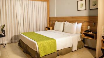 Quarto de hotel com uma cama grande, cabeceira de madeira, mesa de cabeceira com abajur, uma cadeira e uma janela com cortinas.
