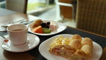Uma mesa com uma xícara de café, um prato de comida incluindo pães e um sanduíche, e um prato com frutas e sobremesa.