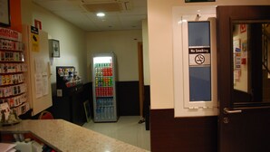 Um saguão de hotel com uma mesa de recepção, uma máquina de venda automática e um aviso de 'Não Fumar'.
