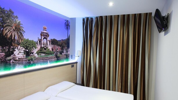 Quarto de hotel com uma cama, uma janela grande com vista para uma fonte e um edifício, e uma televisão fixada na parede.
