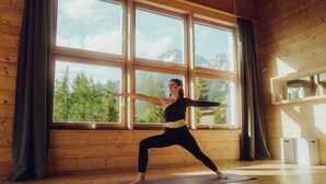 Um tapete de yoga no chão com uma pessoa vestida de preto executando uma postura de yoga em frente a janelas amplas.