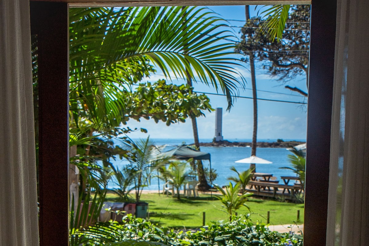 Vista de uma varanda com guarda-corpo de madeira, uma rede pendurada e um jardim verdejante com palmeiras e um corpo d'água ao fundo.