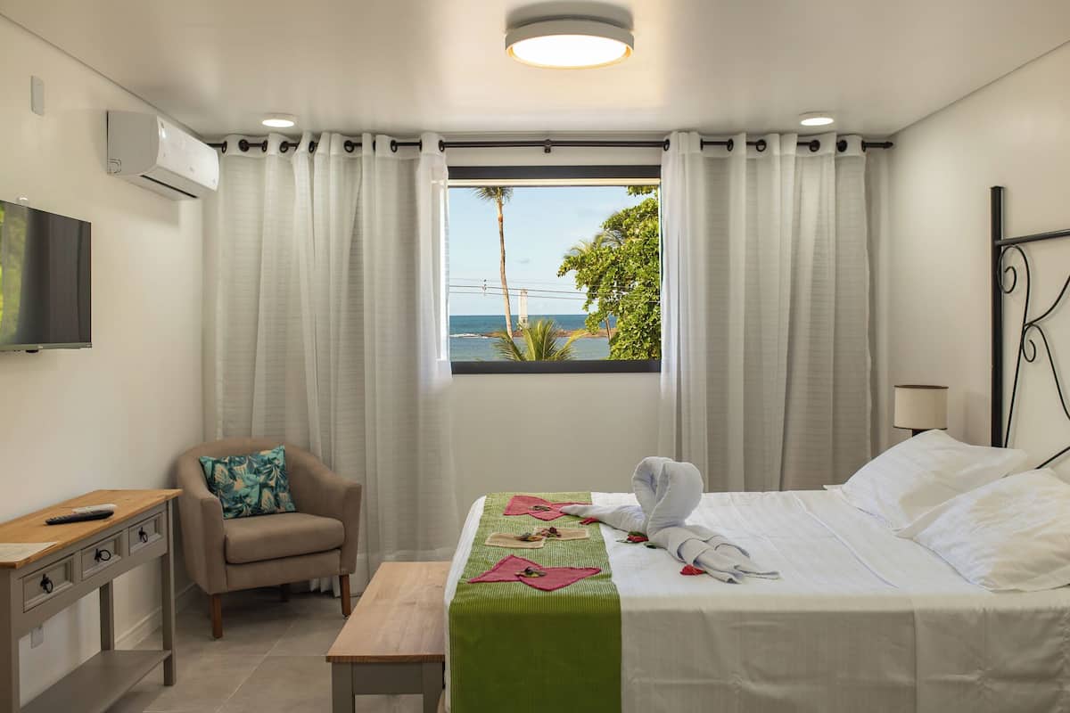 Quarto de hotel com cama, cadeira, escrivaninha, televisão e janela com vista para o mar.