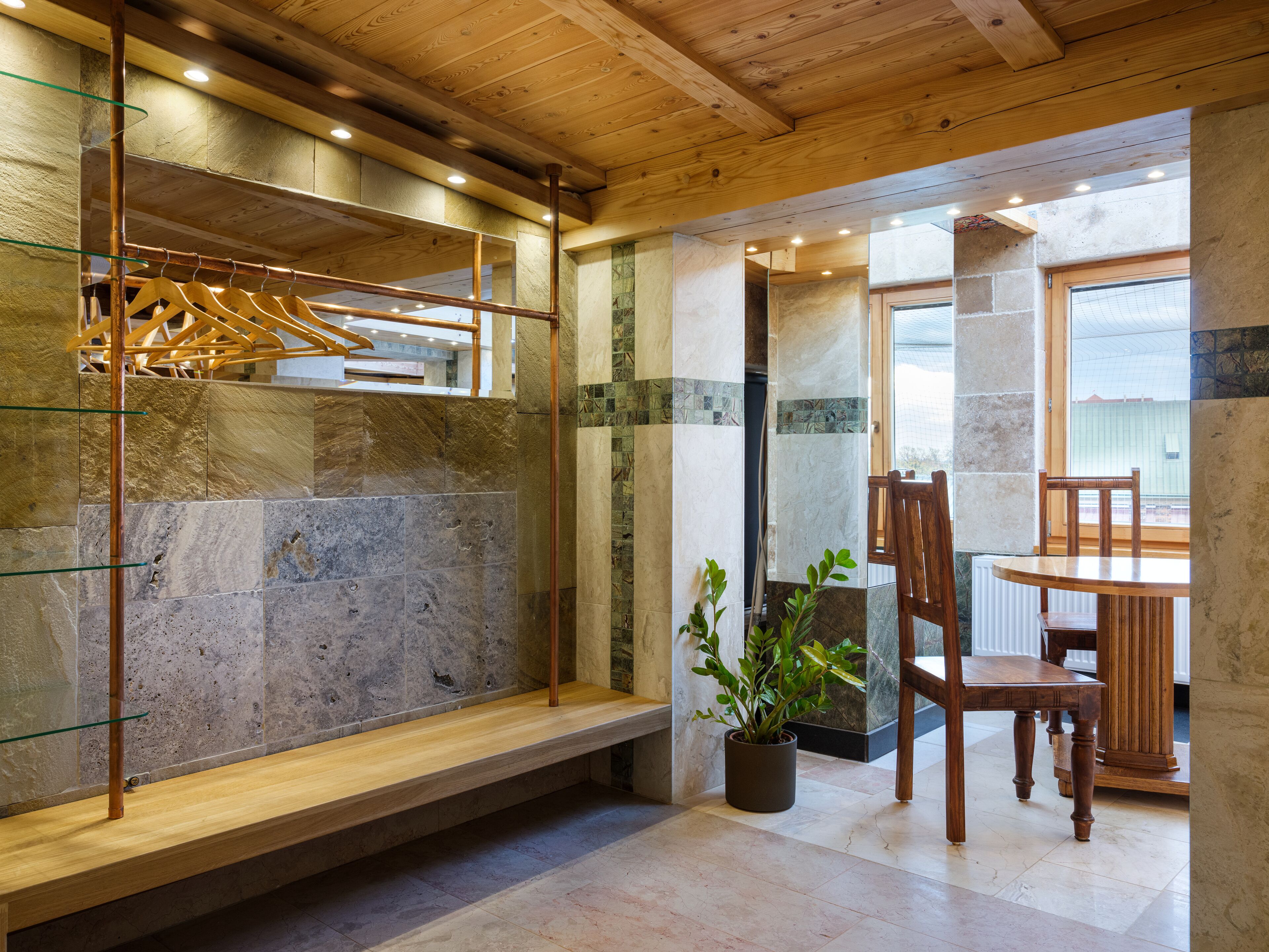 Teto de madeira, um banco, uma mesa com cadeiras e um vaso com planta em um ambiente com paredes de pedra e uma janela.