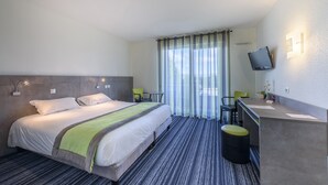 Quarto de hotel com cama, escrivaninha, cadeira, televisão e janela com cortinas.
