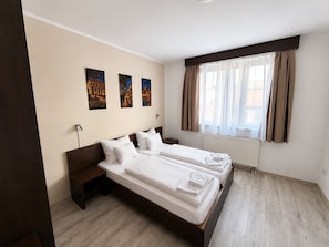 Quarto de hotel com duas camas, uma mesa de cabeceira, janela com cortinas e três quadros pendurados na parede.