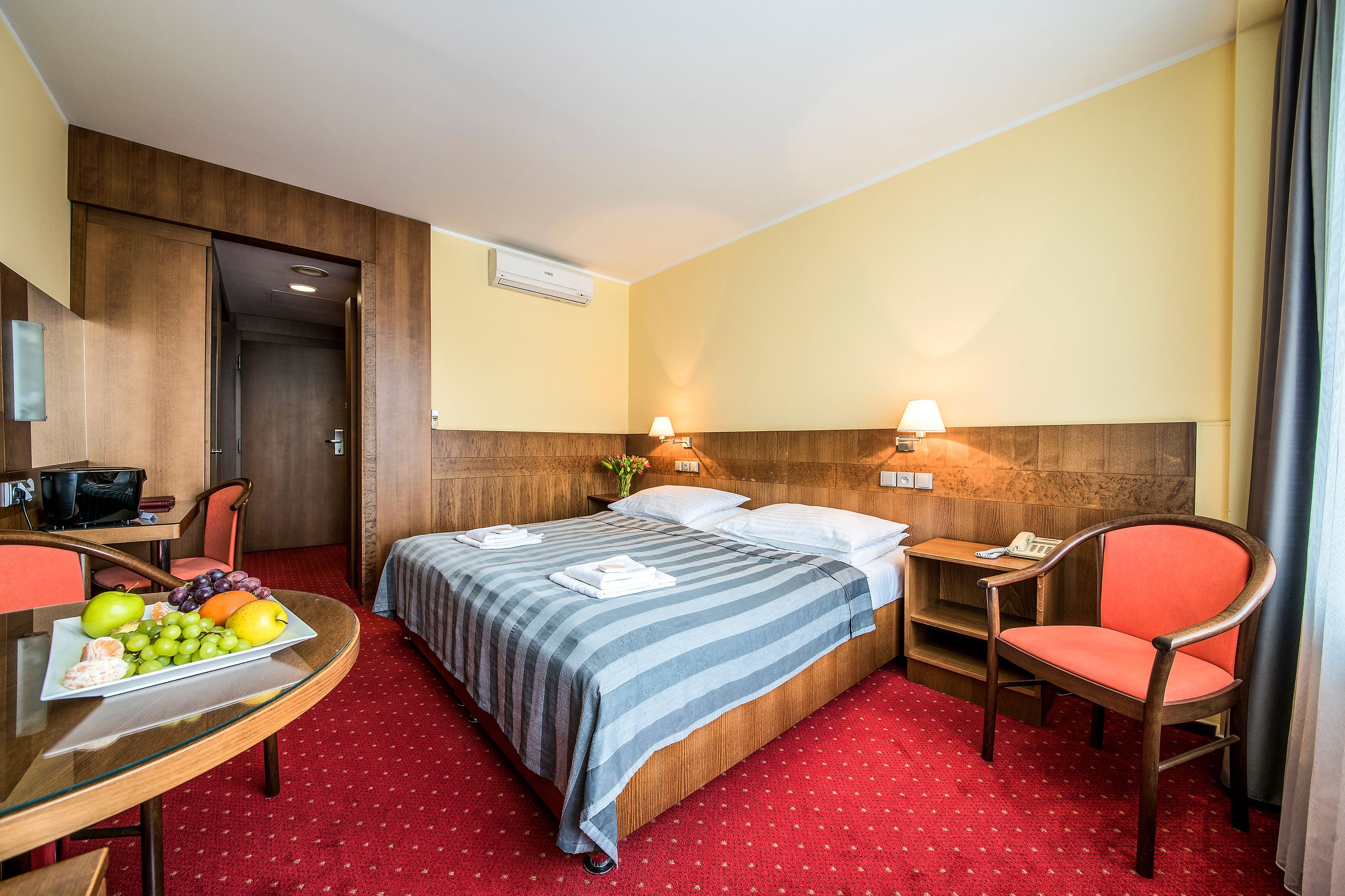 Quarto de hotel com cama, escrivaninha, cadeira e mesa de jantar com frutas.