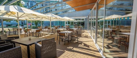 Uma área de pátio ao ar livre com mesas e cadeiras, coberta por guarda-sóis, e janelas amplas com vista para os edifícios ao redor.