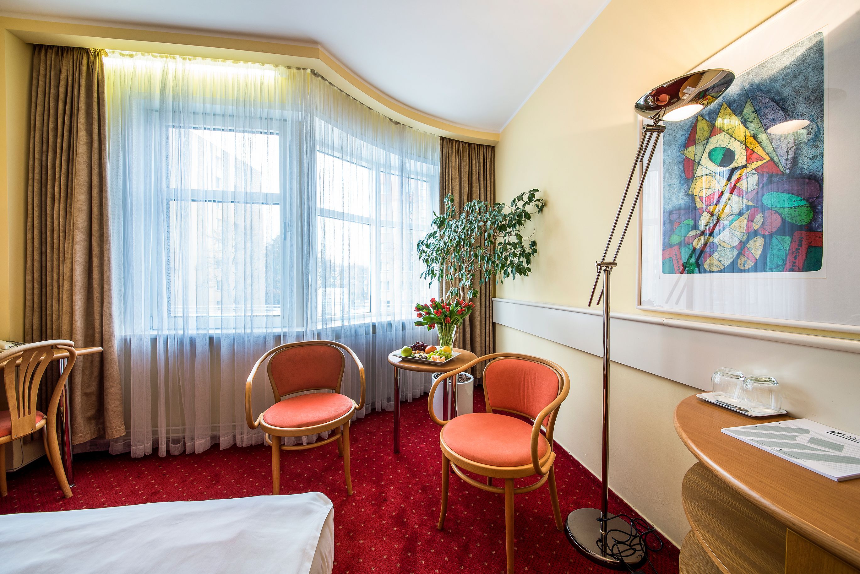 Quarto de hotel com uma cama, duas cadeiras, uma mesa pequena com um vaso de flores, um abajur e um quadro na parede.