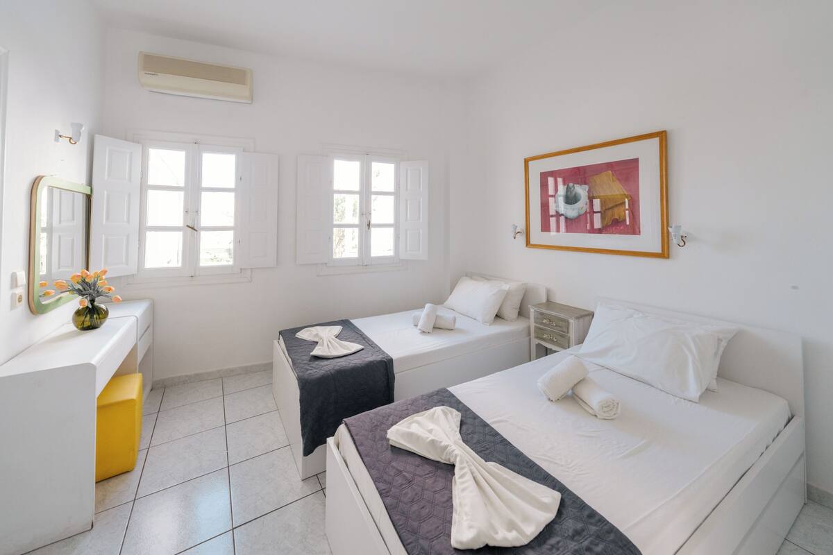 Quarto de hotel com duas camas, banheiro, escrivaninha com espelho e janela.