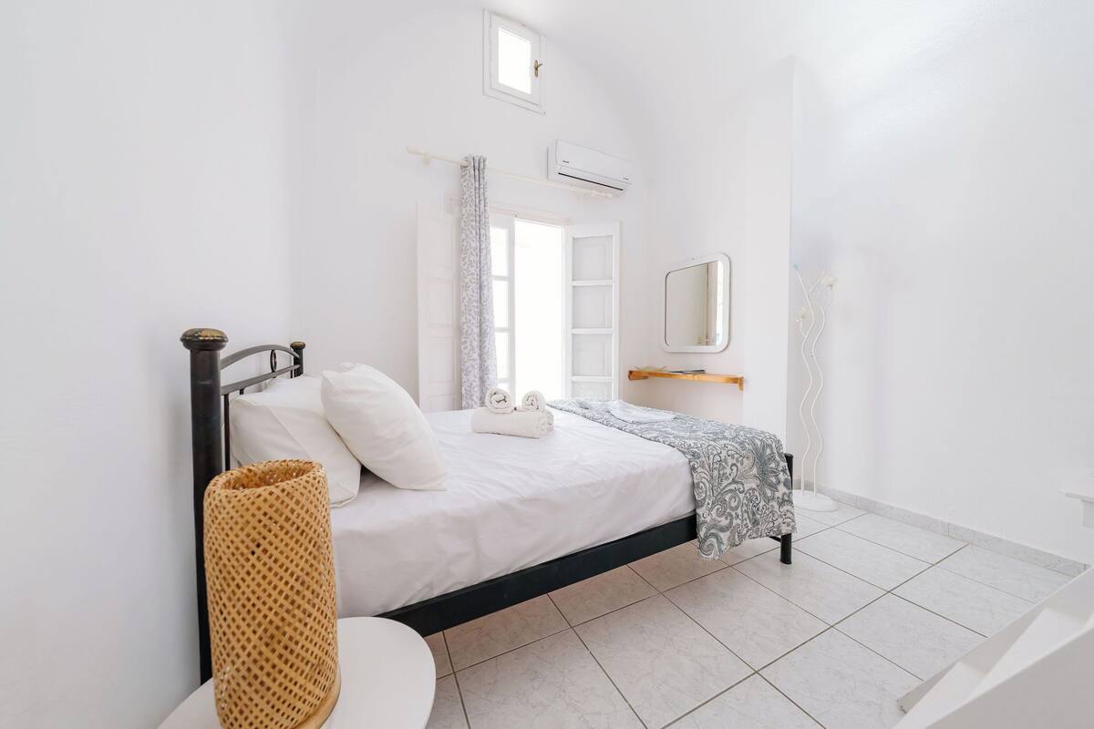 Quarto com cama, mesa de cabeceira, cesto de vime, janela com cortinas, ar condicionado e espelho na parede.