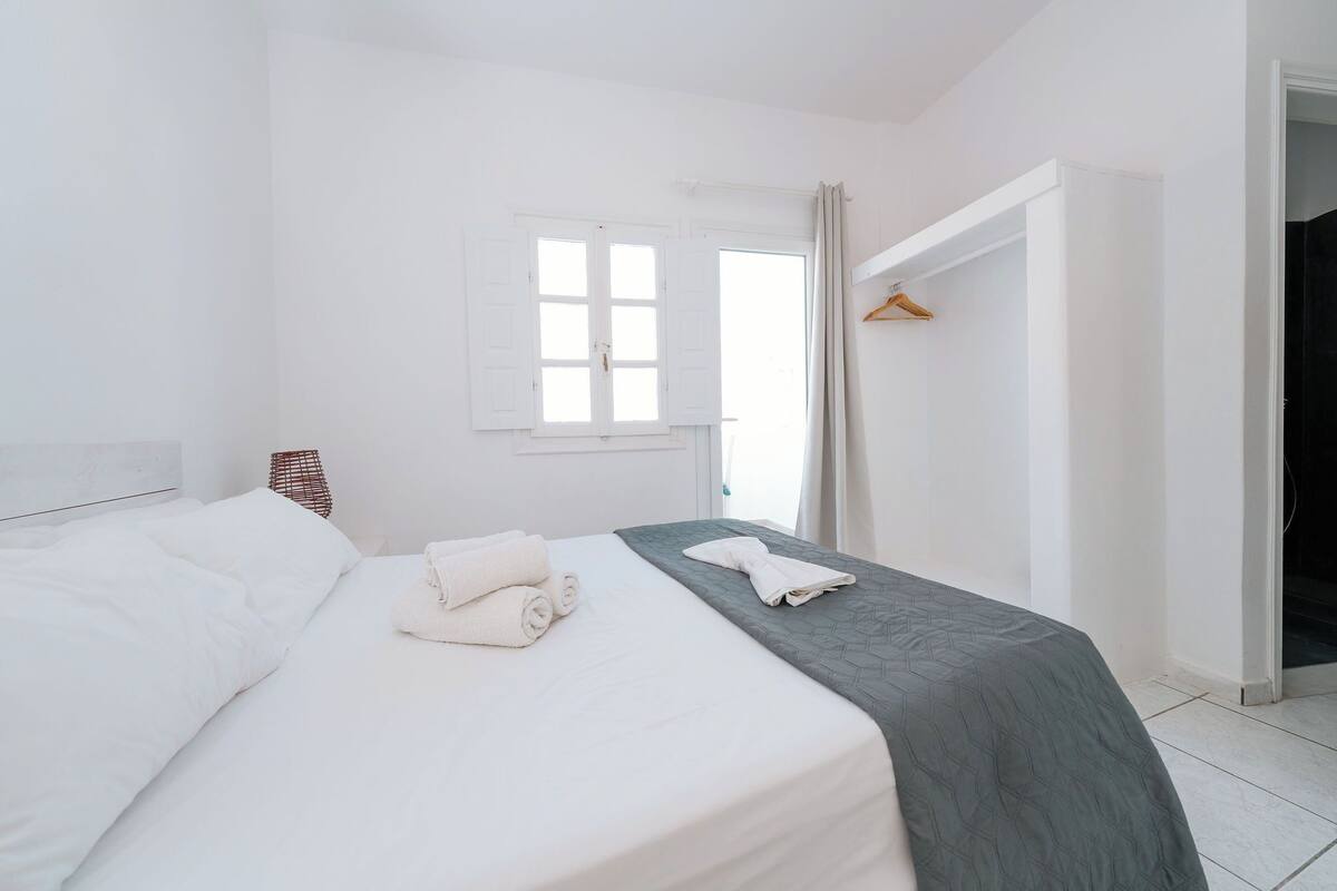 Quarto de hotel com cama, cabideiro e janela com cortinas.