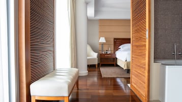 Quarto de hotel com um banco de madeira, uma cama, uma mesa de cabeceira, um abajur e vista para o exterior.