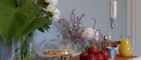 Uma mesa com um vaso de vidro cheio de flores, uma placa de madeira, uma cesta de frutas, um jarro de vidro e uma garrafa de suco.