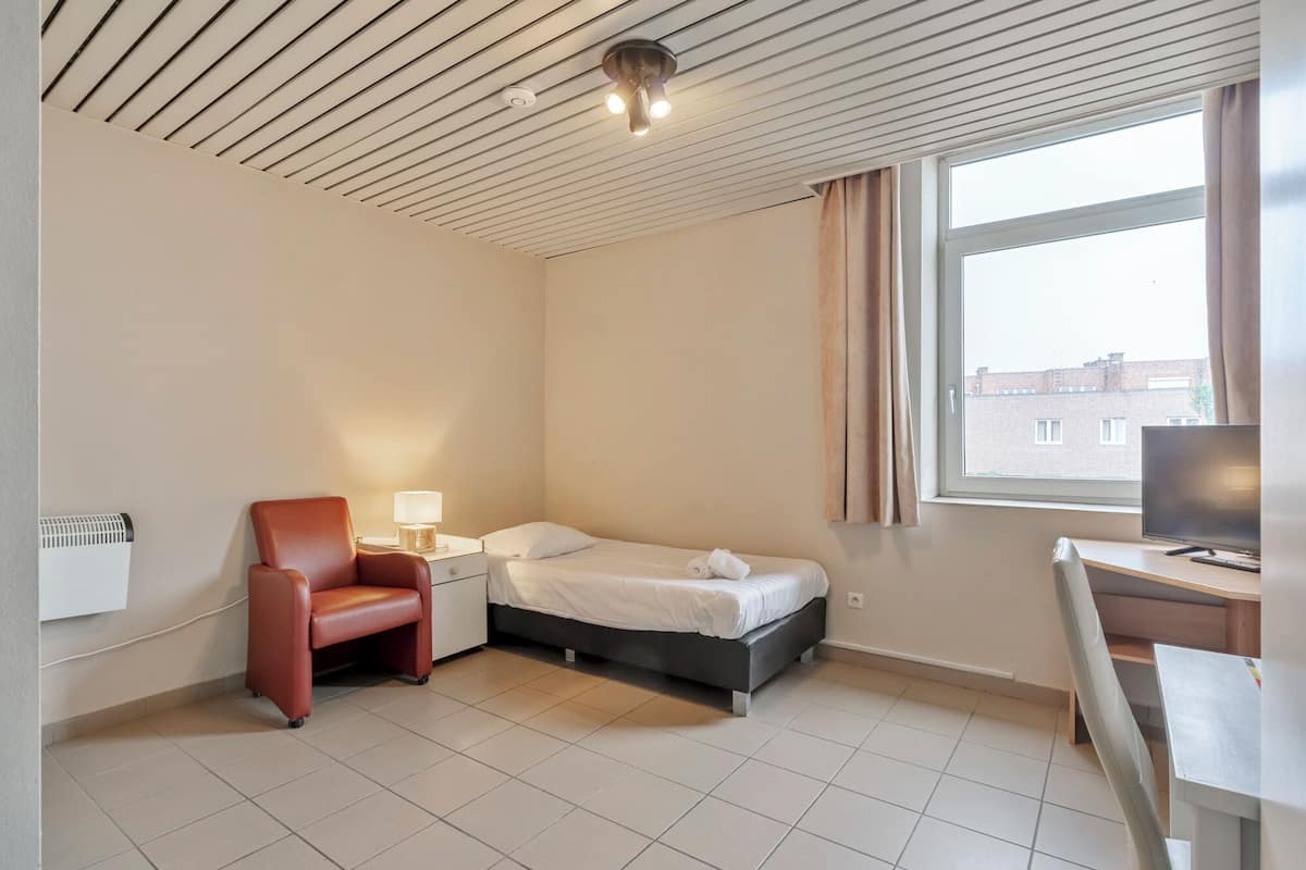Quarto de hotel com cama, escrivaninha, cadeira, abajur, radiador e janela com vista para edifícios.