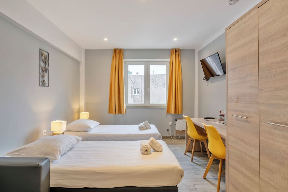 Quarto de hotel com duas camas, uma escrivaninha, uma cadeira, uma televisão e um guarda-roupa.