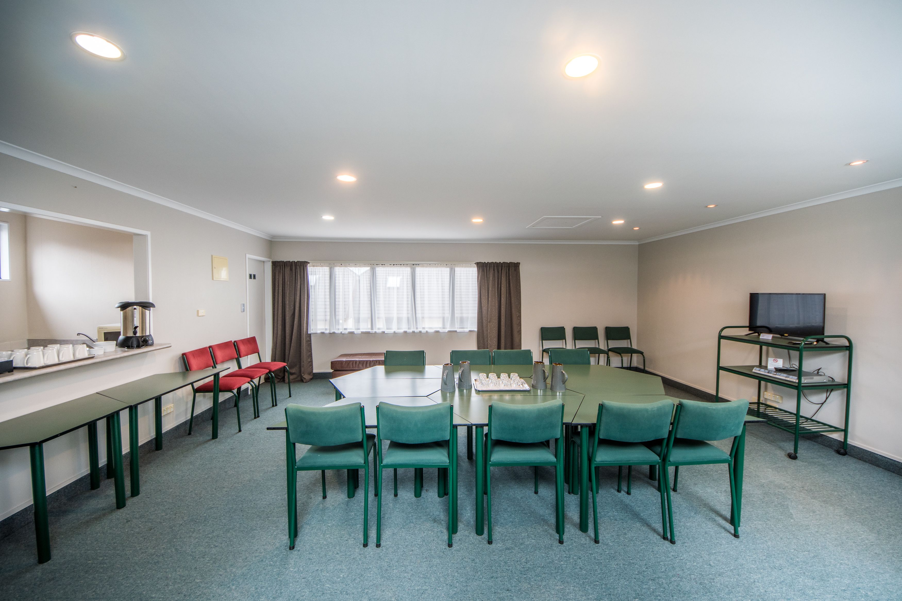 Uma sala de conferência com uma mesa comprida, cadeiras verdes, uma televisão em um suporte e uma janela com cortinas.