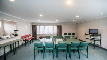 Uma sala de conferência com uma mesa comprida, cadeiras verdes, uma televisão em um suporte e uma janela com cortinas.