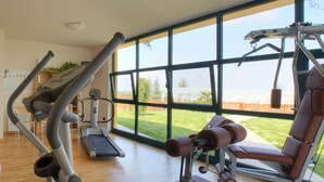 Uma academia com aparelhos de cardio, um banco de musculação e amplas janelas que dão para um jardim.